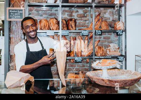 Smiling man working in a bakery taking a piece of cake with a tongs ...