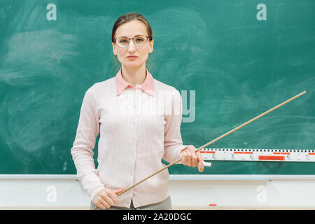 strict teacher standing in front of blackboard in class with a pointer ...