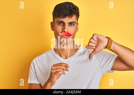 Young indian man holding fanny mustache standing over isolated yellow ...