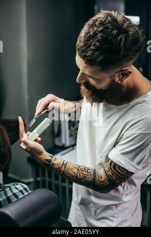 Serious tattooed barber cutting hair of young man with piercings ...