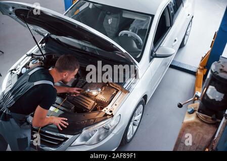 Uses lighting equipment. Man in work uniform repairs white automobile ...
