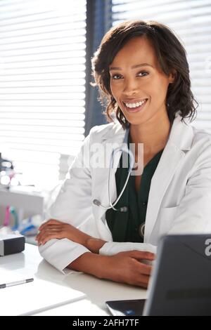 Female Doctor Or GP Wearing White Coat Sitting At Desk In Office Using ...
