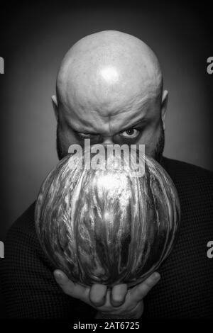 A bald man holds a bowling ball in front of his face squinting at the ...