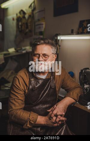 positive, bearded shoemaker in leather apron looking at camera while ...