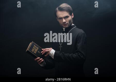 confident catholic priest holding holy bible and looking at camera on ...