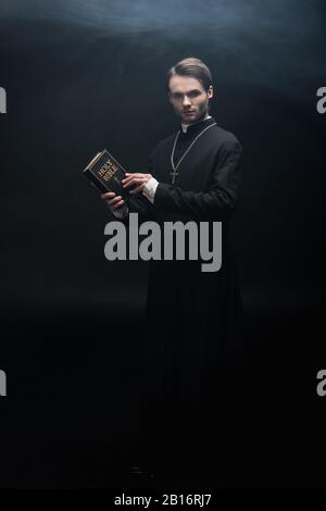 confident catholic priest holding holy bible and looking at camera on ...
