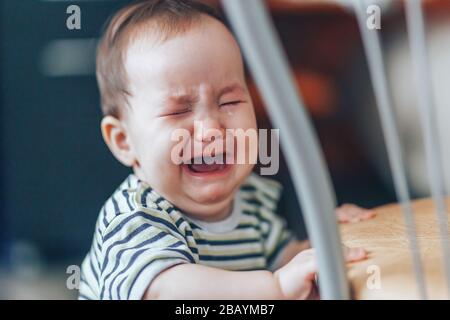Crying toddler baby boy stands holding his mother legs in the kitchen ...