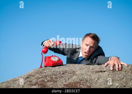 Desperate businessman dangling off a rock cliff making an urgent call ...