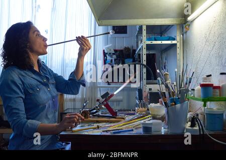Young skilled female lampworking master blowing into long tube while ...