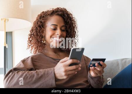 Young woman smiling, holding a credit card and smartphone for online ...