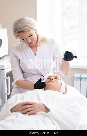 Cosmetologist in black gloves applying a hydrating facial mist spray on ...
