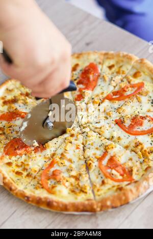 Pizza cutter wheel slicing ham pizza with capsicum and olives on wooden ...