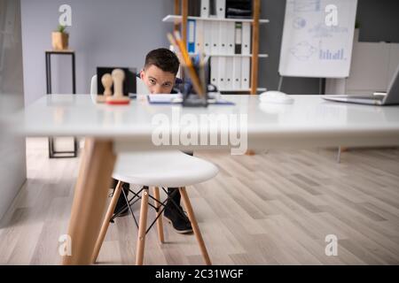 Frightened Young Businessman Hiding Behind Chair At Workplace Stock ...