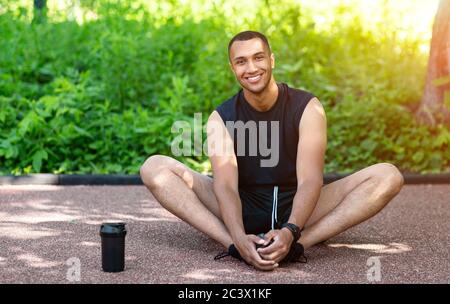 Happy millennial guy doing flexibility exercises after jogging at park ...
