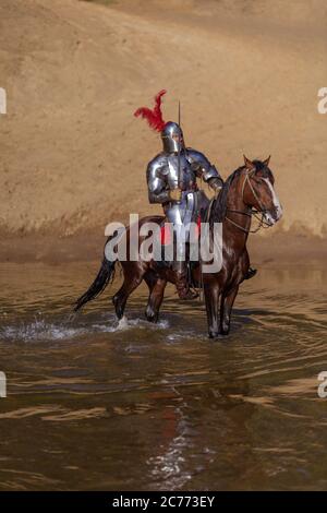 A young adult man in knightly armor rides a horse on a river along a ...