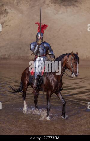 A young adult man in knightly armor rides a horse on a river along a ...