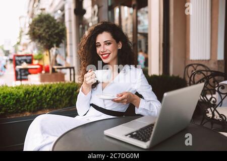 joyful curly girl holding coffee cup and using laptop in bed during ...