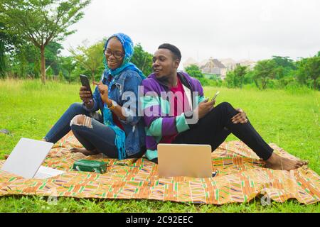 Happy Nigerian couple using the cellphones while on a picnic on a grass ...
