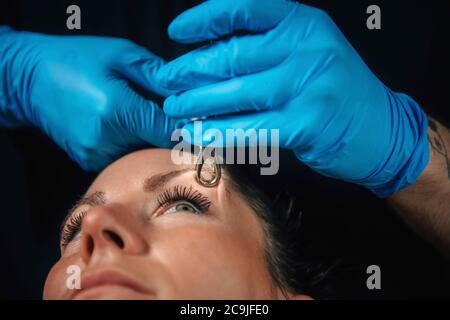 Young woman getting her eyebrow pierced, body piercer wearing blue ...