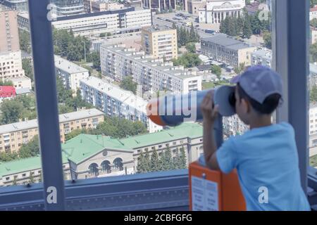 The boy looks into a telescope from a panoramic window of a high-rise ...