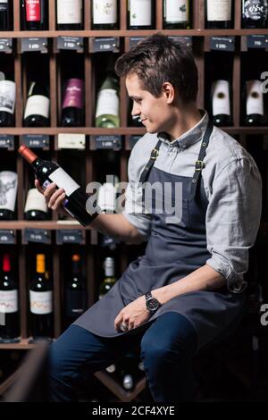Focused young male sommelier in suite looking at red wine in bottle ...
