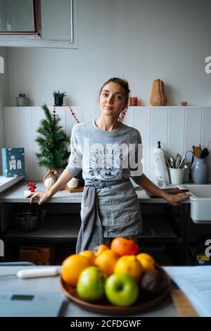 Positive housewife leaning on a kitchen set with vegetables while ...