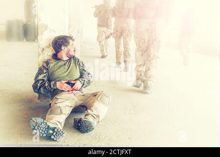 Wounded on stomach soldier, sitting at floor, squeezing bleeding wound ...