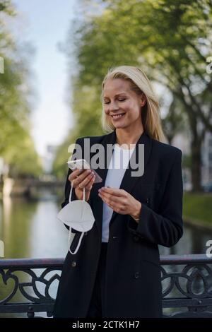 Smiling businesswoman with mobile phone and protective face mask ...