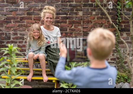 Boy photographing smiling mother and sister through smart phone in ...