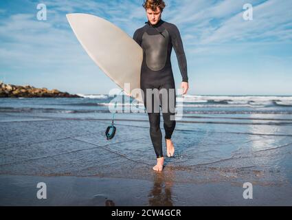 Portrait of young surfer leaving the water with surfboard under his arm ...