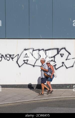 A couple of holidaymakers walking past a wall sprayed with ugly ...