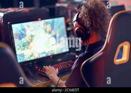 Rear view of female gamer girl with headset sits in a dark room behind ...