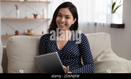 Smiling Asian woman using digital tablet near window at home for remote ...