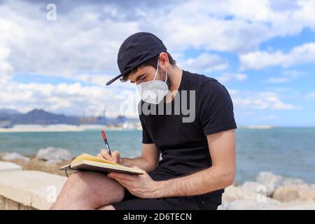 Male author in face mask and cap sitting with crossed legs on fence ...