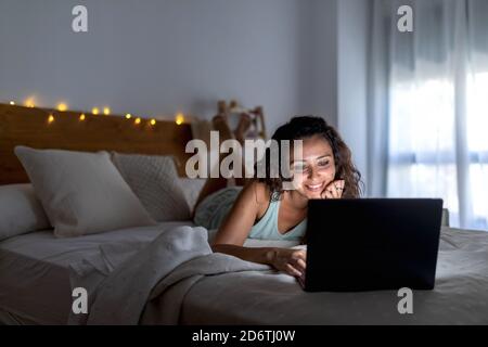 Delighted female in nightwear lying on bed in dark room and watching ...