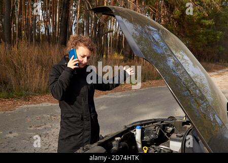 An upset woman stands near her broken car and talking on a cell phone on the road in autumn ...