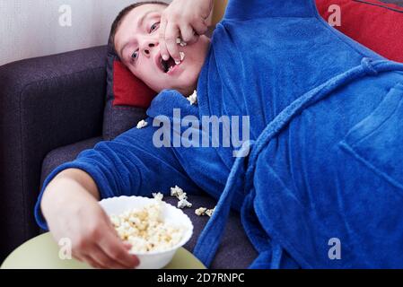 Fat man in a robe eating popcorn while lying on the couch, obesity and ...