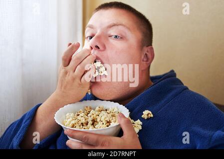Fat man in a robe eating popcorn while lying on the couch, obesity and ...