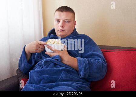 Fat man in a robe eating popcorn while lying on the couch, obesity and ...