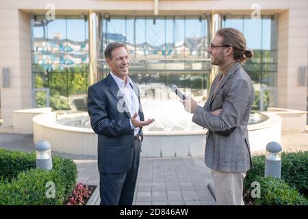 Long-haired male journalist having interview with smiling mature male in suit in front of camera ...