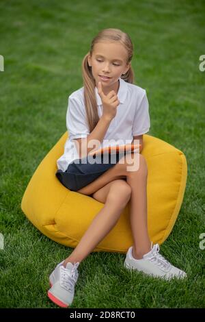 Cute fair-haired schoolgirl sitting on a bag chair and showing her tablet Stock Photo - Alamy