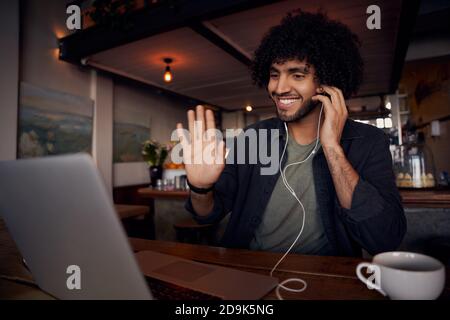 Happy man in earphones waving hand while taking video call on laptop at ...