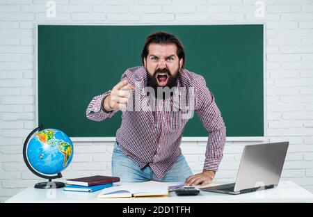 Angry Bearded teacher in classroom sits at desk. Back to school ...