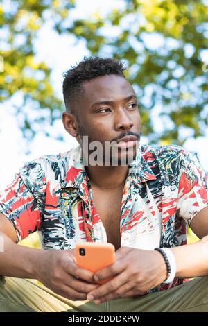 Thoughtful young man with smart phone looking out of window at home ...