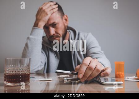 Male junkie sitting at the table with drugs and syringe, grunge room ...