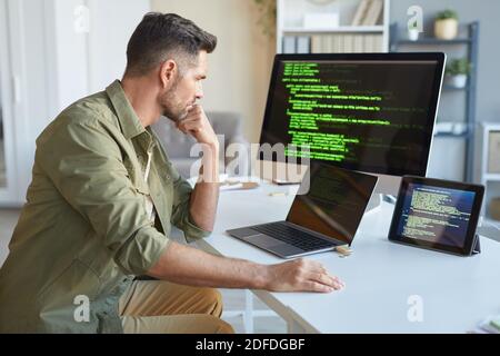Serious mature software developer looking at code on computer screen while decoding data and ...