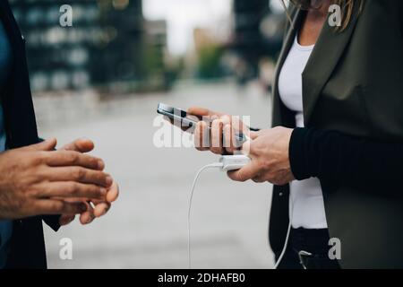 Midsection of mature businesswoman charging mobile phone while standing ...