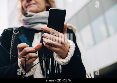 Midsection of businesswoman charging mobile phone while standing with ...
