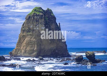 An immense rock formation in wavy seawater under a majestic cloudscape ...