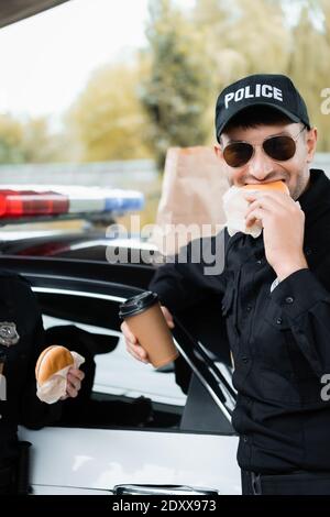 Policewoman eating burger and holding coffee to go near colleague and ...